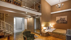 Um quarto amplo com uma cama grande, um espaço de estar com uma poltrona e uma mesa, uma escada que leva a um andar superior e um candelabro.