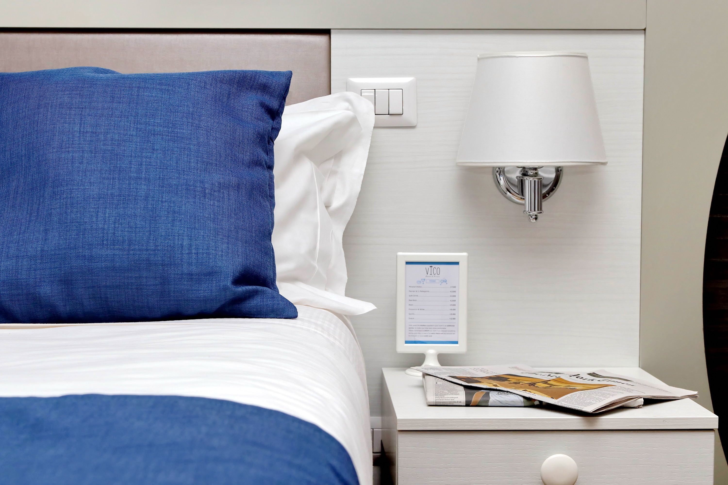 Uma cama bem arrumada com roupa de cama azul e branca, um abajur, uma mesinha de cabeceira com um jornal e um interruptor de luz fixado na parede.