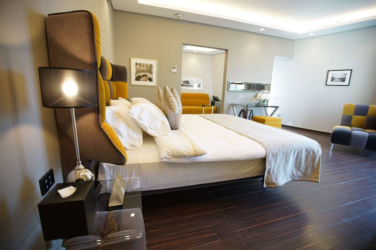 Quarto de hotel com uma cama grande, uma mesa de cabeceira com luminária, uma mesinha com caixa de lenços e uma cadeira.