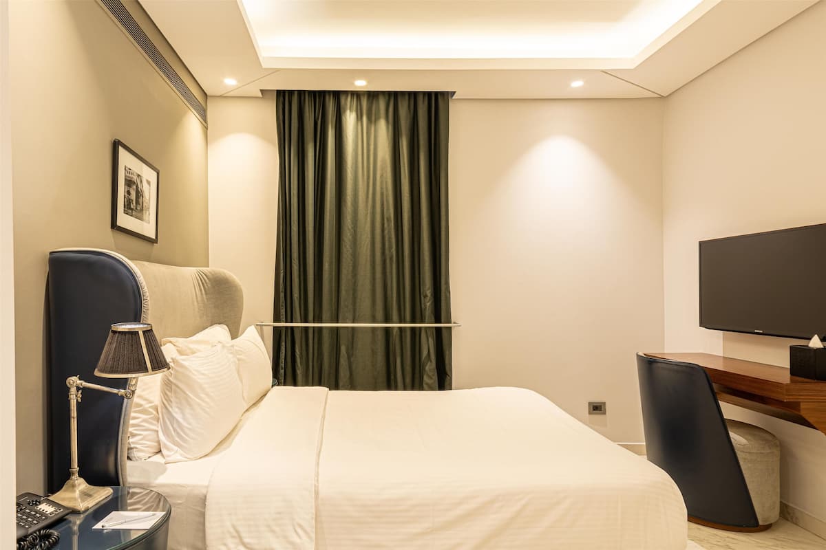 Quarto de hotel com cama, escrivaninha com cadeira, televisão, luminária e telefone.