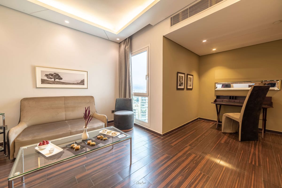 Uma sala de estar moderna com um sofá, uma mesa de centro de vidro com petiscos, uma poltrona e uma escrivaninha com cadeira.