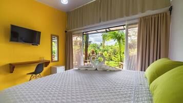 Quarto de hotel com cama, televisão na parede, escrivaninha com cadeira e janela com cortinas.