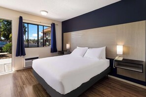 Quarto de hotel moderno com uma cama grande, mesa de cabeceira com abajur, prateleira embutida e uma janela ampla com cortinas.