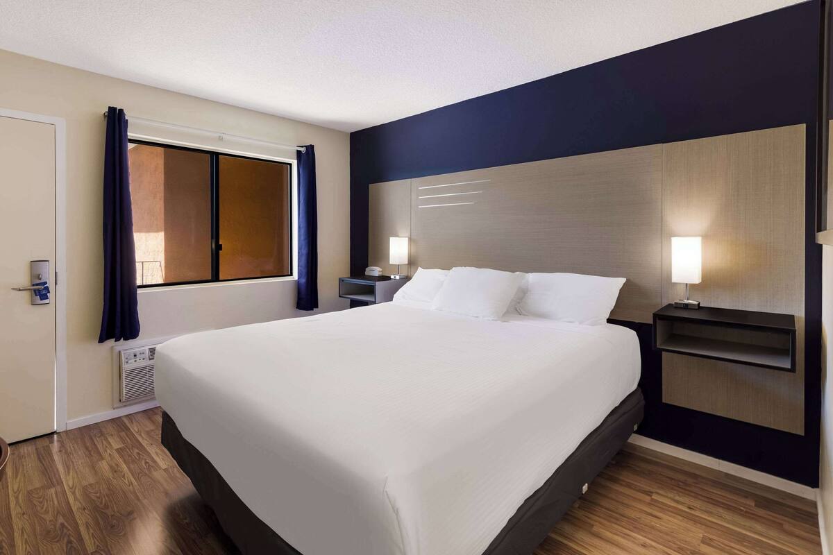 Quarto de hotel com uma cama grande, mesa de cabeceira com abajur, guarda-roupa e janela com cortinas.