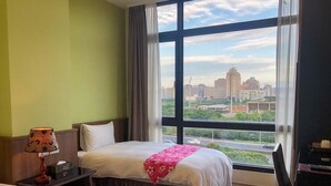 Quarto de hotel com uma grande janela, uma cama com colcha floral rosa, uma mesa de cabeceira com abajur e vista para a cidade.