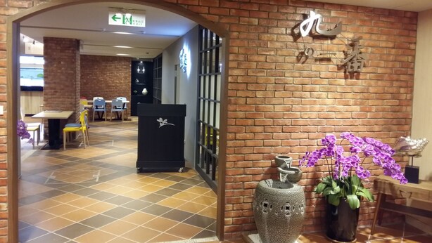 Um hall de hotel com parede de tijolos, balcão de recepção, lixeira preta e planta em vaso com flores roxas.