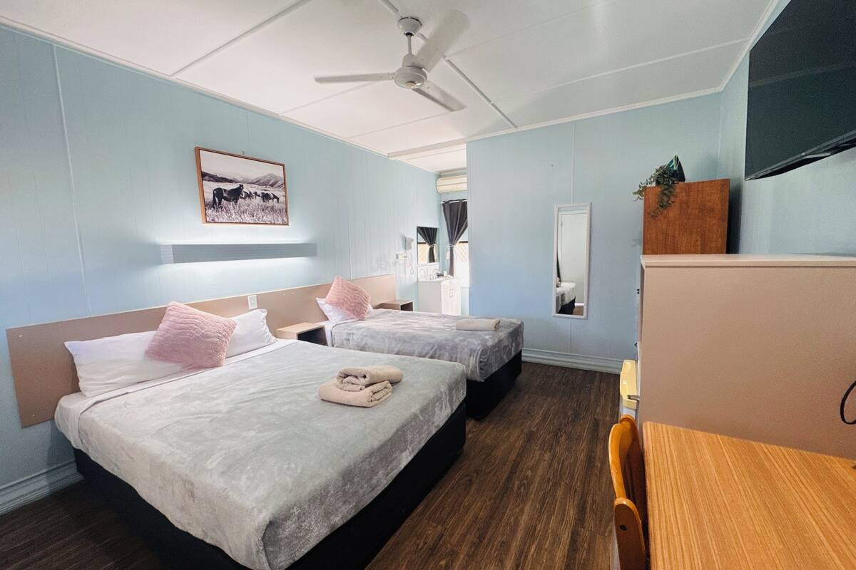 Quarto de hotel com duas camas, um ventilador de teto, uma mesa de madeira, um espelho e um quadro na parede.