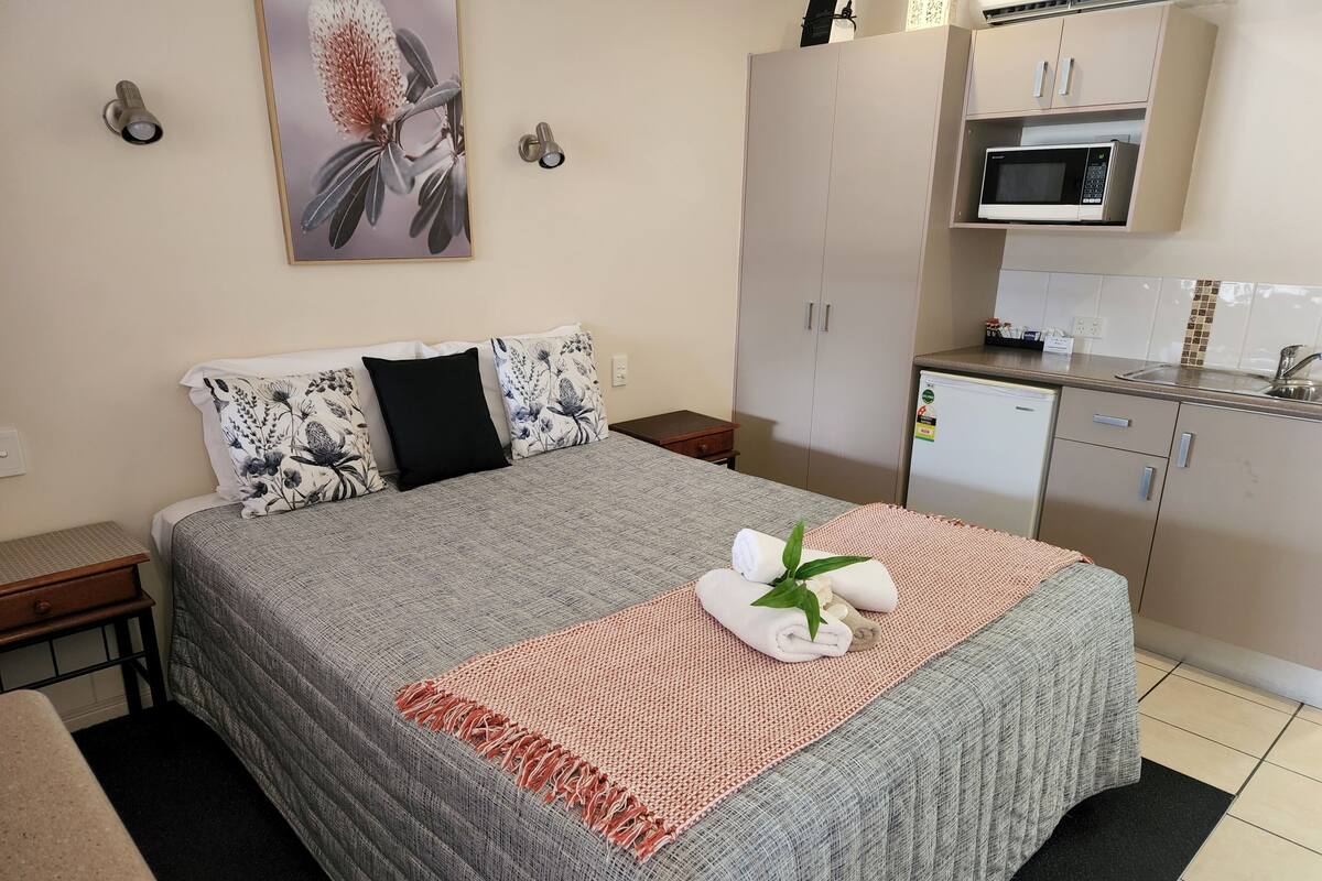 Cama bem arrumada com colcha cinza, manta rosa e branca, e arranjo de toalhas. Há uma pequena mesa de cabeceira, luminária fixada na parede e uma obra de arte floral emoldurada sobre a cama.