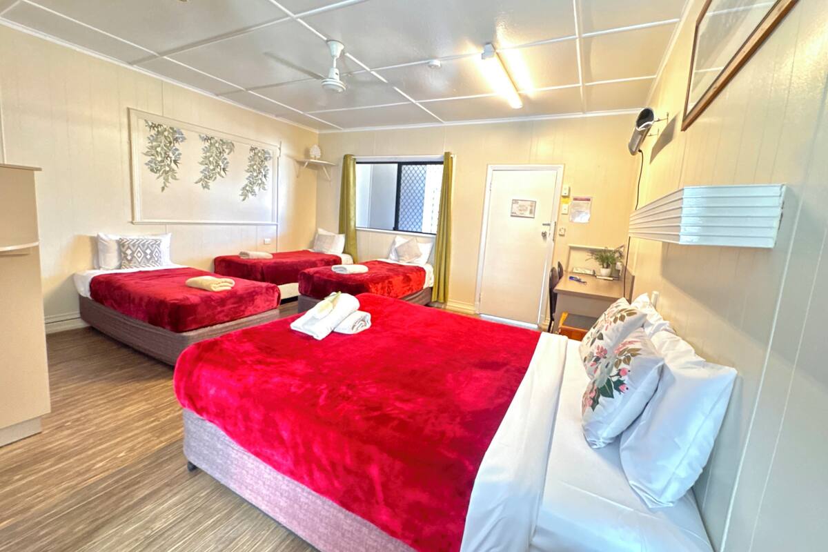 Quarto de hotel com duas camas, uma escrivaninha, uma cadeira, uma janela com cortinas e um ventilador de teto.