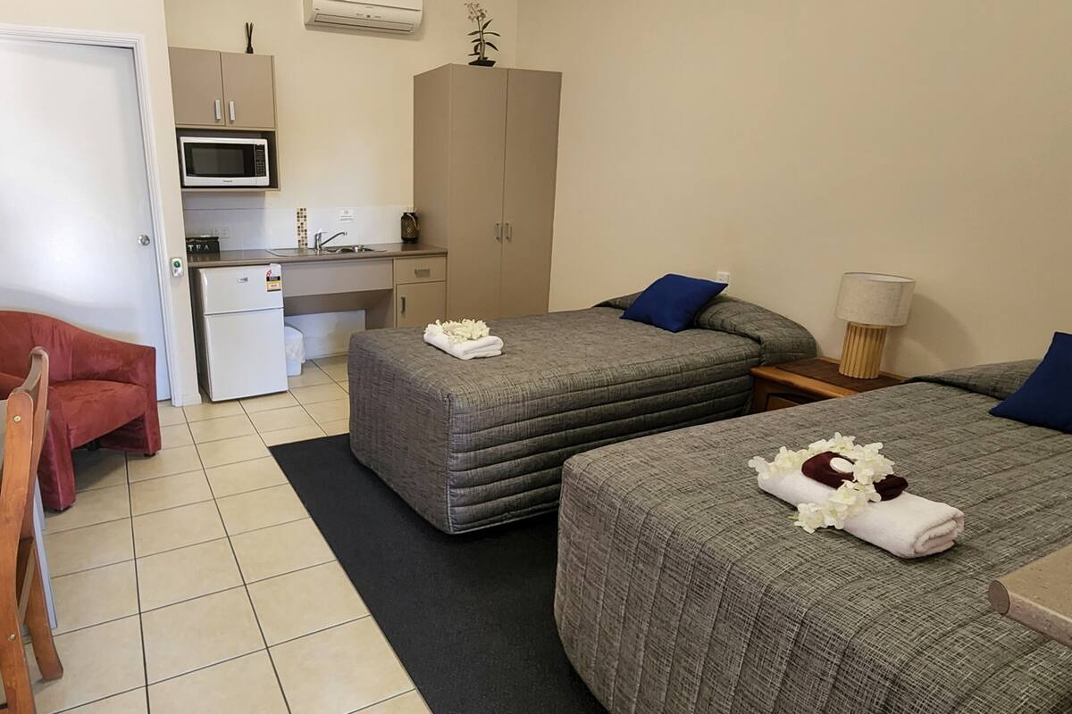 Quarto de hotel compacto com cozinha americana, duas camas com toalhas, uma cadeira vermelha e uma escrivaninha.