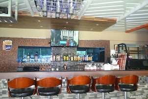 Uma área de bar com balcão, diversas garrafas, copos e uma tela de televisão. Quatro cadeiras de madeira estão dispostas em frente ao bar.