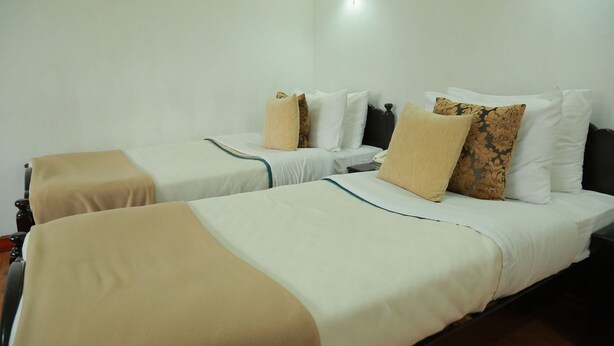 Quarto de hotel com duas camas, cada uma com roupa de cama branca e almofadas decorativas bege. Há uma luminária fixada na parede acima das camas.