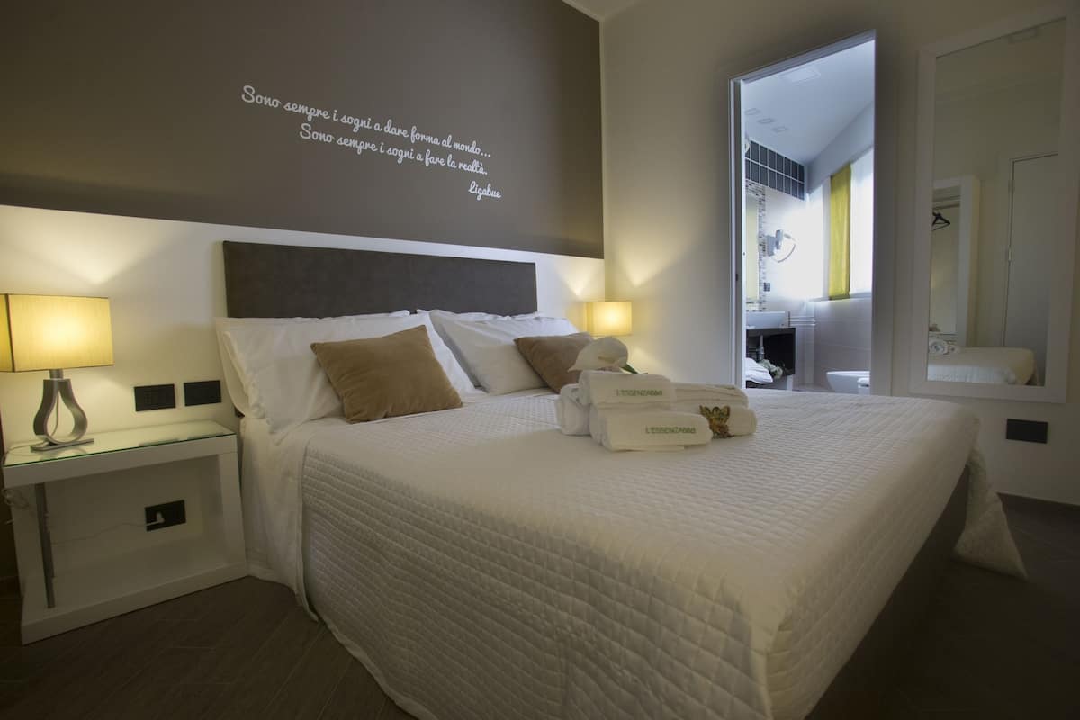 Quarto de hotel com cama grande, duas luminárias de cabeceira, uma frase decorativa na parede e vista para o banheiro.