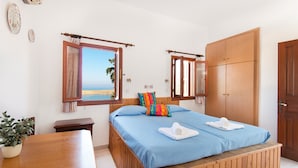 Quarto com cama de madeira, escrivaninha de madeira, planta em vaso e vista para o mar pela janela.