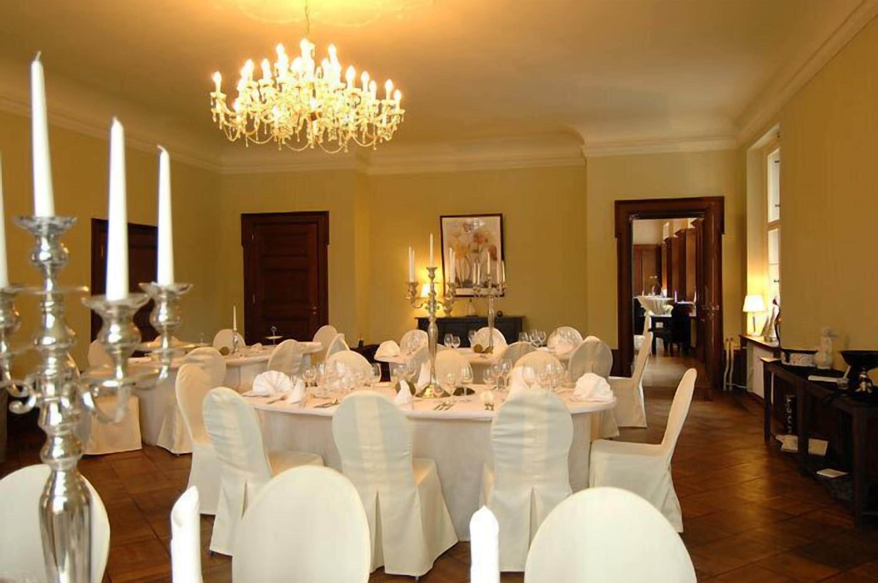 Uma sala de jantar formal com mesas redondas, cadeiras brancas e um candelabro.
