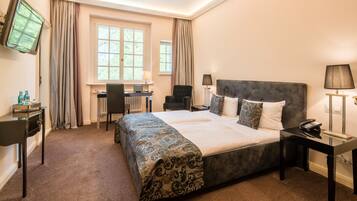 Quarto de hotel com uma cama grande, uma escrivaninha, uma cadeira e uma televisão na parede.