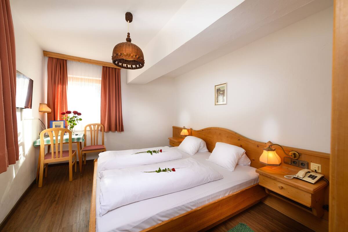 Quarto de hotel com cama de madeira, duas mesas de cabeceira, escrivaninha com cadeira e uma janela grande com cortinas.