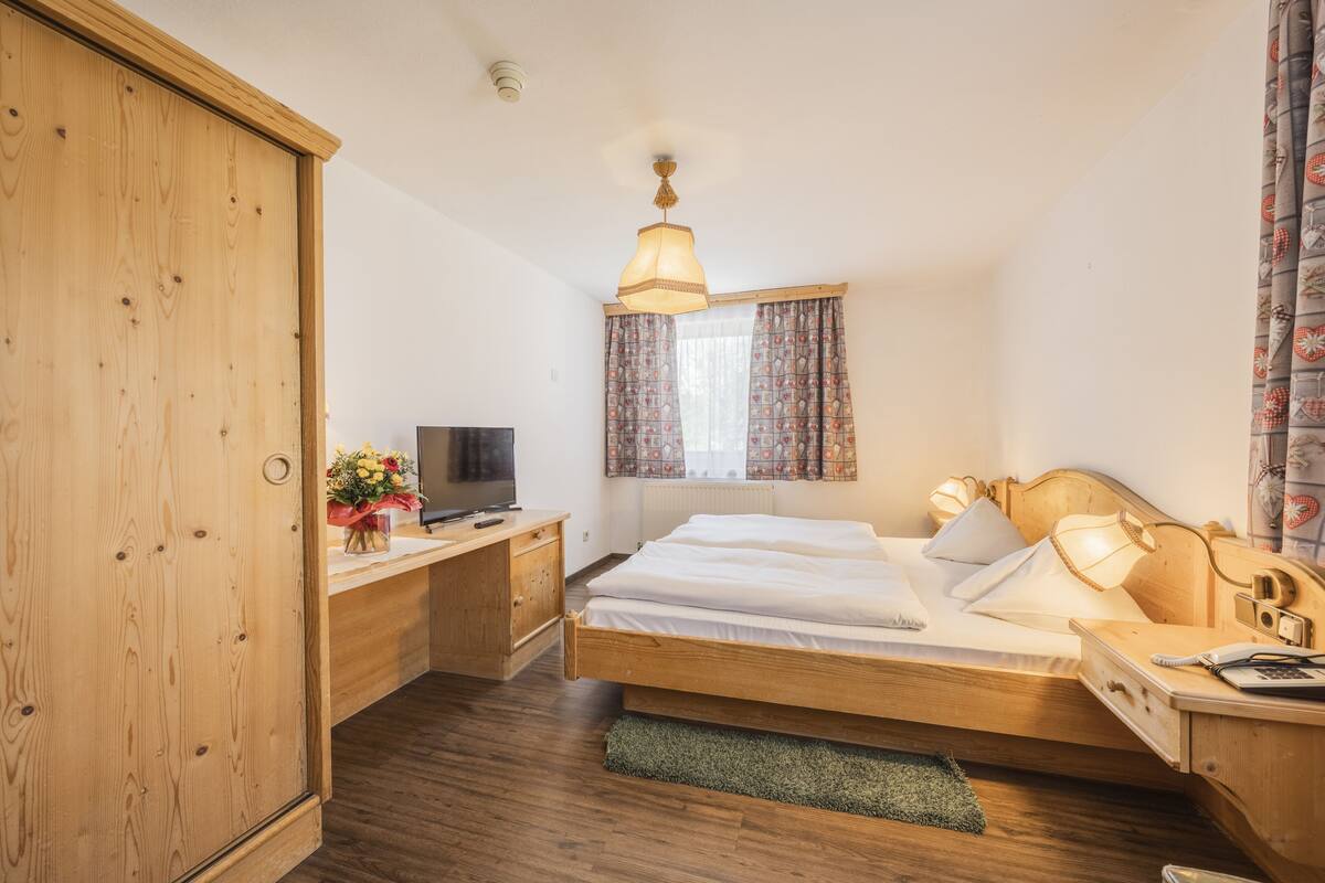 Quarto com cama de madeira, escrivaninha com computador, guarda-roupa e janela com cortinas.
