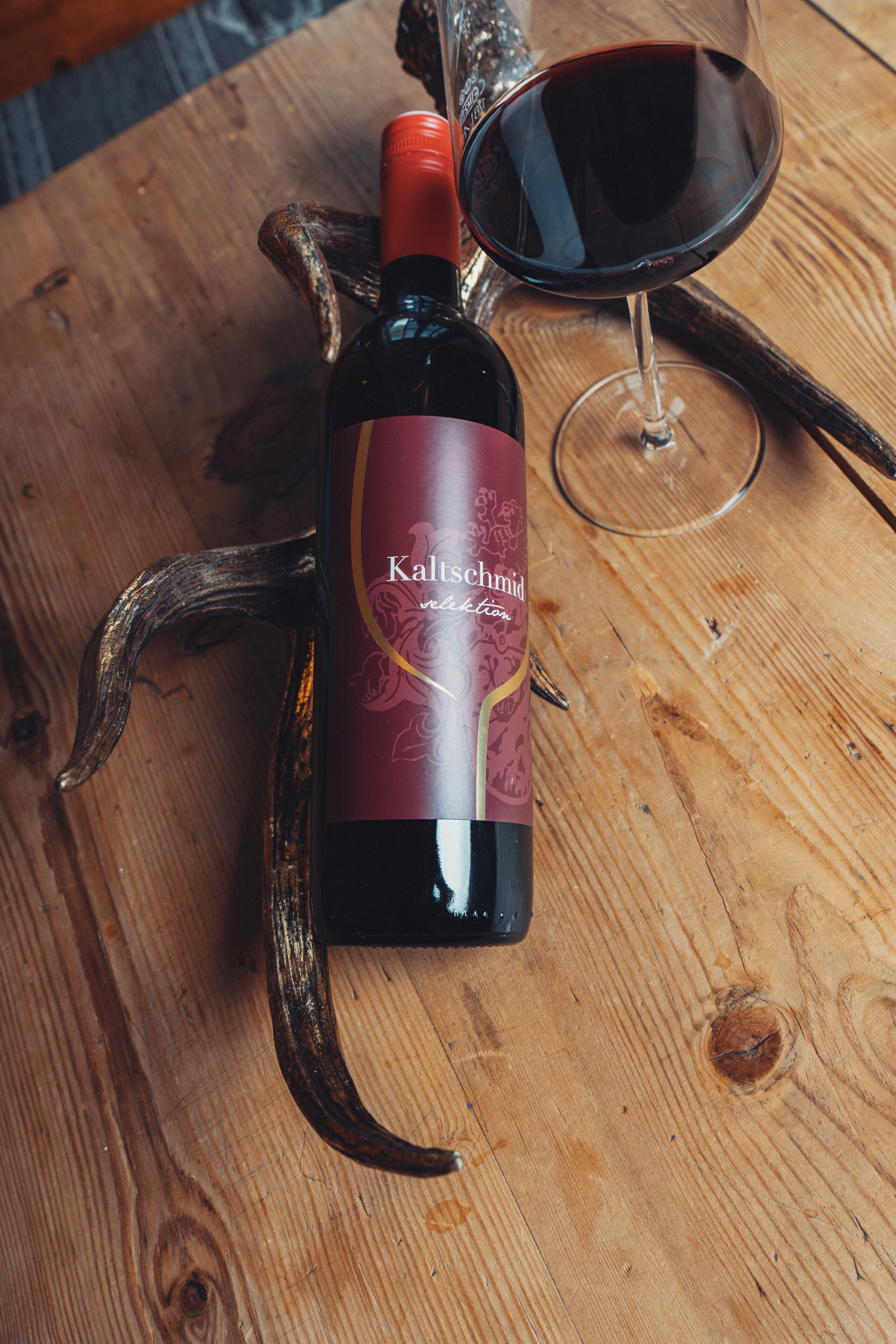 Uma garrafa de vinho tinto Kaltschmidt sobre uma mesa de madeira, com uma taça de vinho e uma colher decorativa.