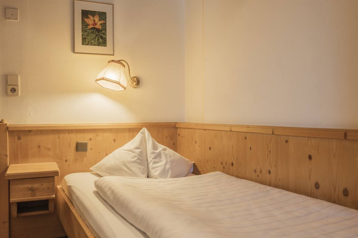 Uma cama de madeira com roupa de cama branca, uma mesa de cabeceira com gaveta, uma luminária fixada na parede e um quadro emoldurado.