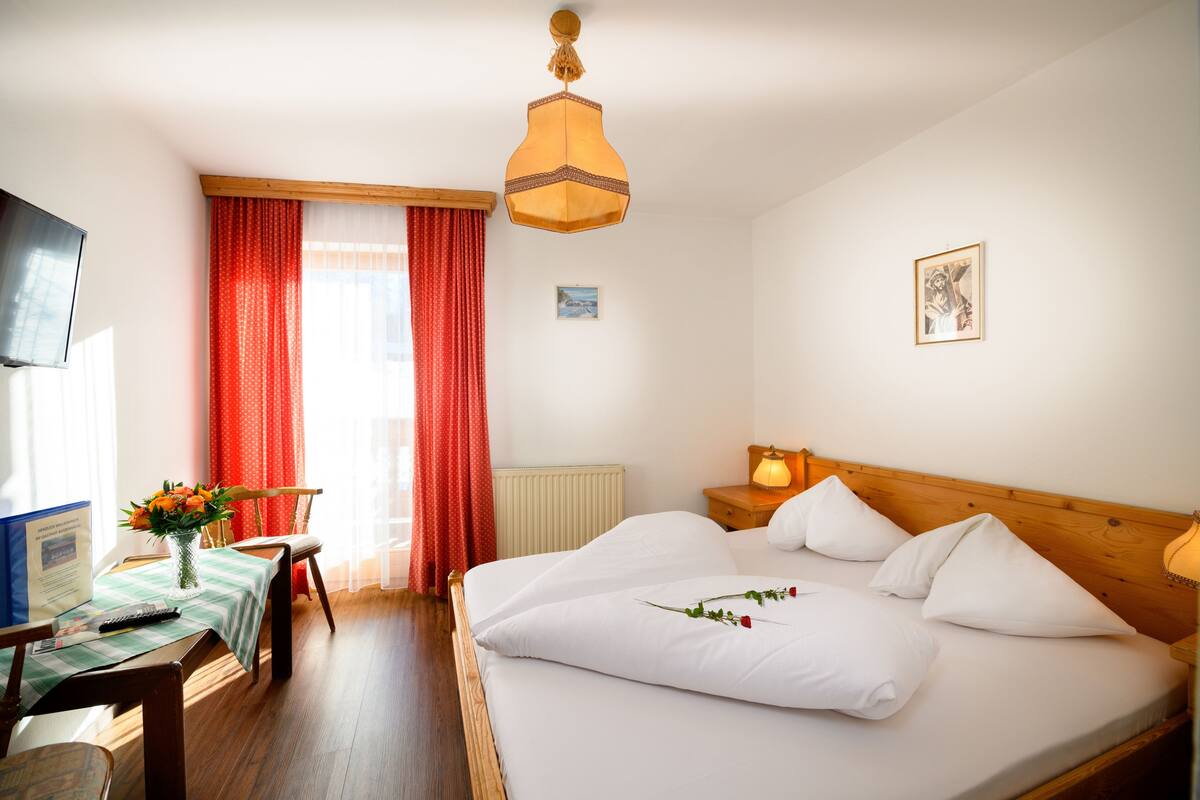 Quarto de hotel com cama, escrivaninha com computador, cadeira, janela com cortinas, TV na parede e um quadro.