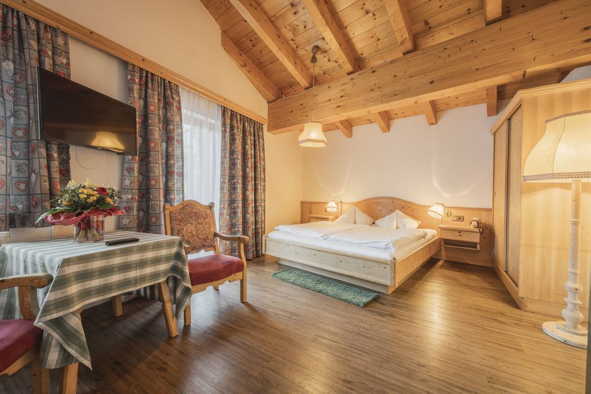 Um quarto aconchegante com teto de madeira, uma cama com mesa de cabeceira, uma mesa com cadeiras e uma TV fixada na parede.