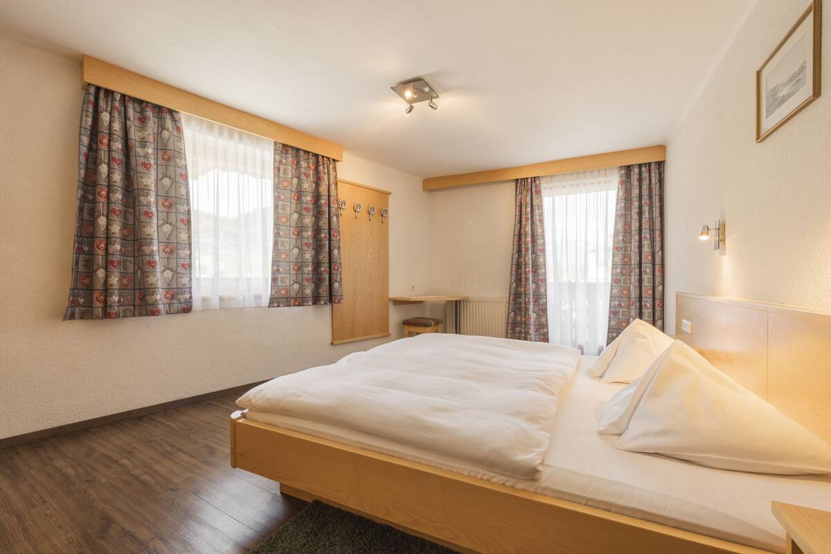 Quarto com cama de madeira, duas janelas com cortinas estampadas, mesa de cabeceira e cabeceira.