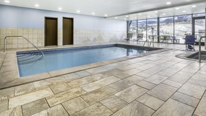 Piscina interna com chão em azulejo, duas portas e vista para montanhas através de amplas janelas.