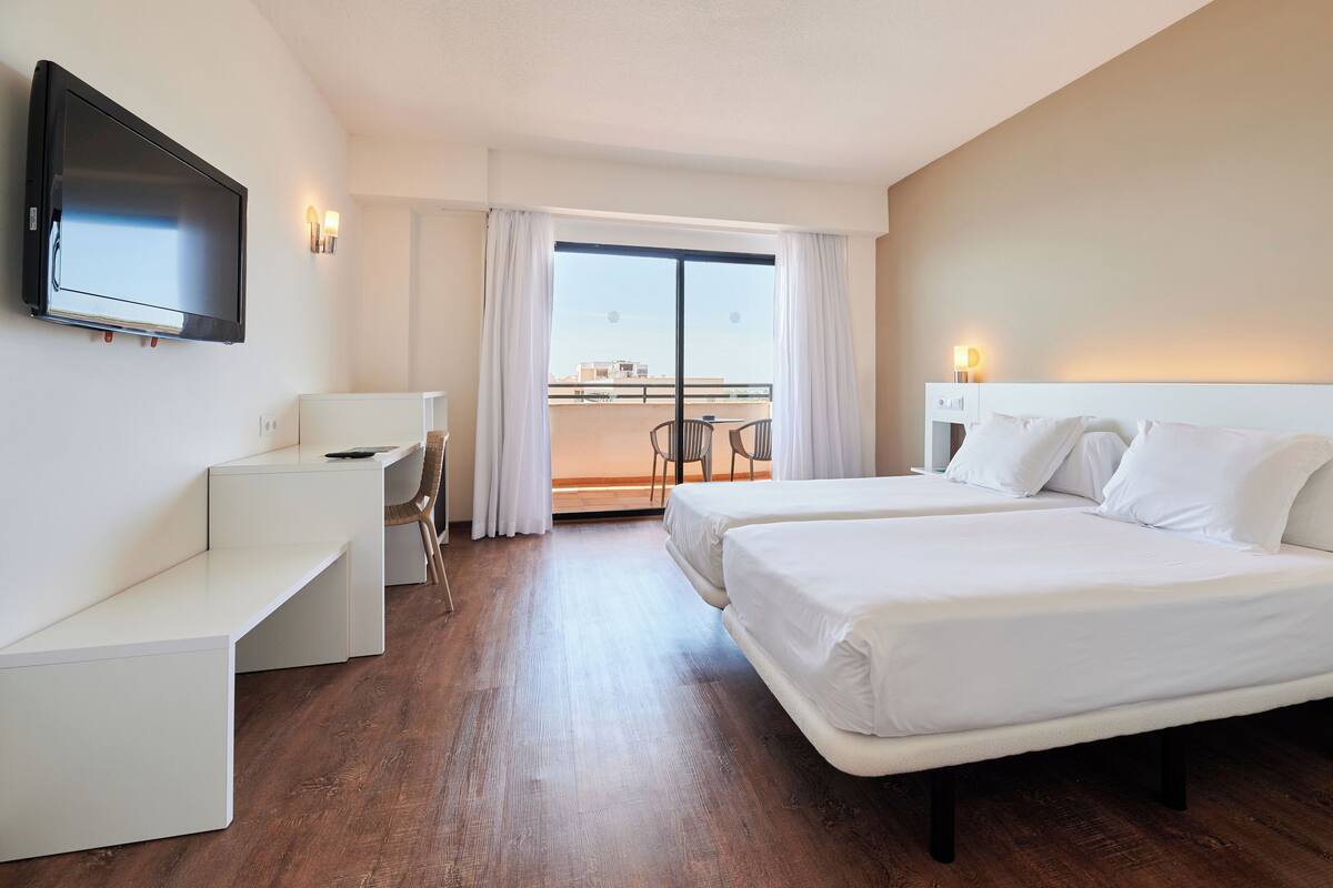 Quarto de hotel com uma cama grande, uma escrivaninha, uma cadeira, TV na parede e varanda com vista.