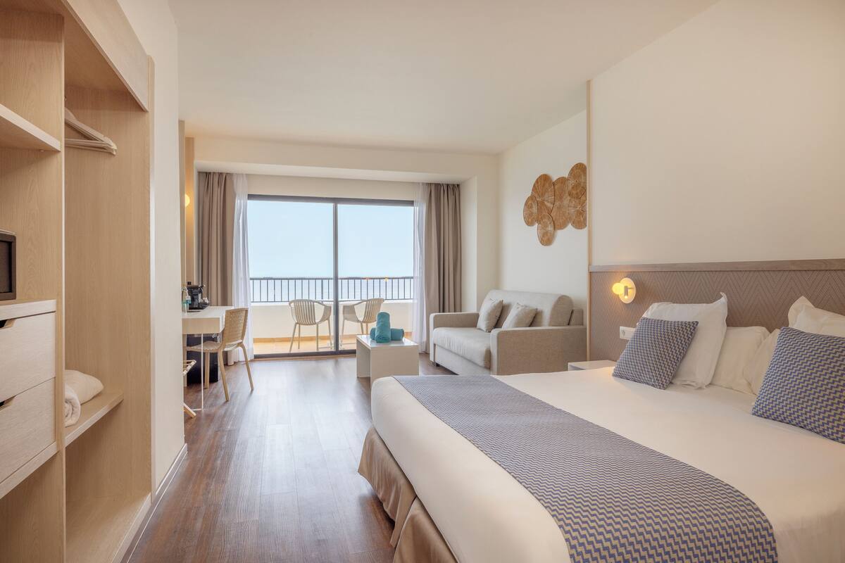 Quarto de hotel com uma cama grande, um sofá, uma mesa de trabalho com cadeiras e uma varanda com vista.