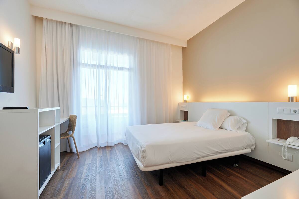 Quarto de hotel com cama, escrivaninha, cadeira, televisão e telefone ao lado da cama.