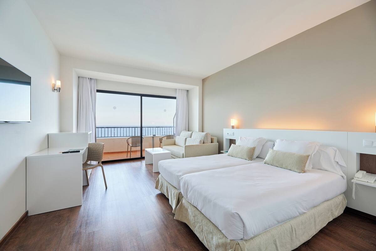 Quarto de hotel com duas camas, uma escrivaninha, uma cadeira e uma varanda com vista.