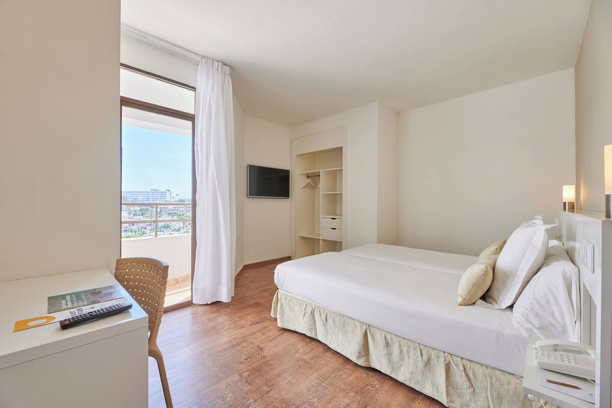 Quarto de hotel com cama, escrivaninha, cadeira, televisão e janela com cortinas.