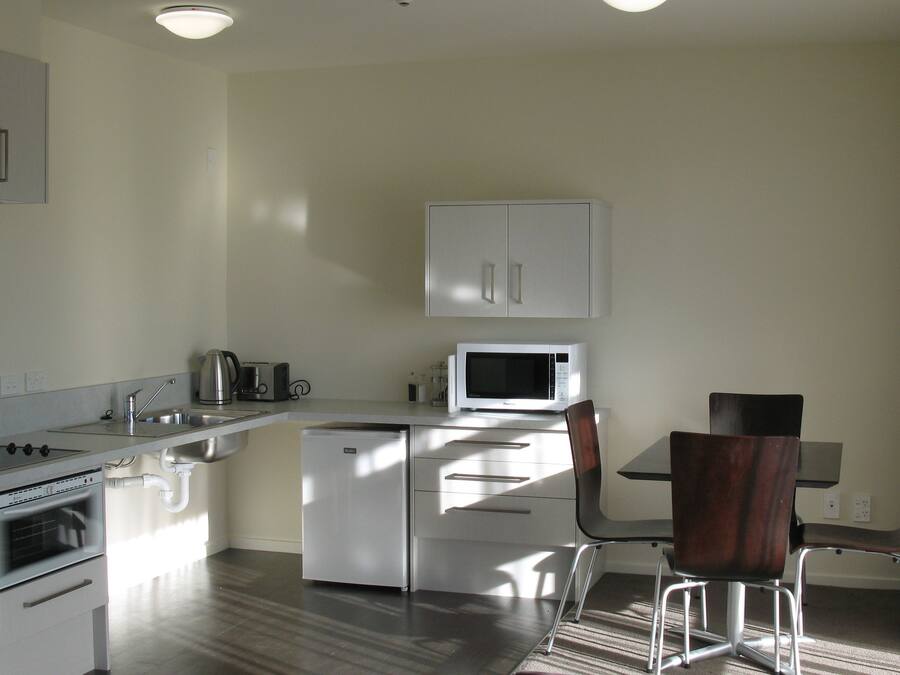 Cozinha compacta com micro-ondas, torradeira e chaleira. Há uma área de refeições com mesa e cadeiras.