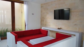 Uma sala de estar moderna com um sofá branco e almofadas vermelhas, uma televisão de tela plana presa a uma parede de pedra, e uma porta de vidro deslizante que dá para uma varanda.