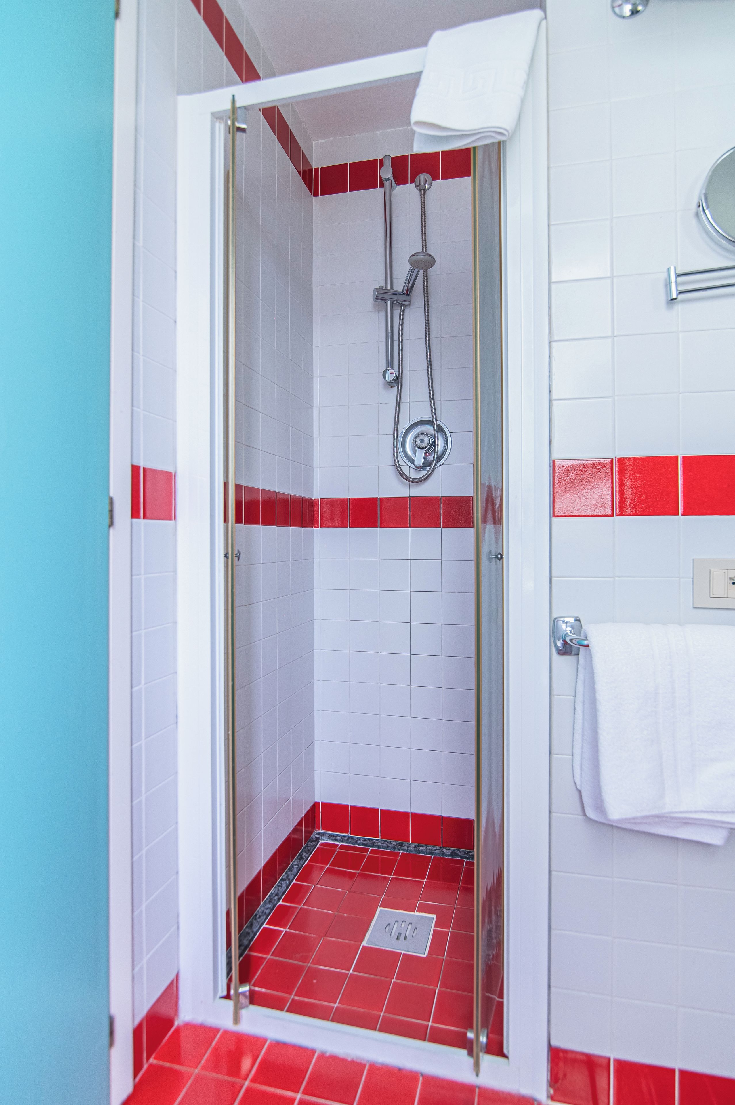 Chuveiro com porta de vidro, toalha branca na parede e paredes com azulejos vermelhos e brancos.