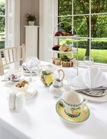 Uma mesa posta para um chá, com um suporte para sobremesas em camadas, saleiro e açucareiro, uma chaleira e um jarro com padrão floral.