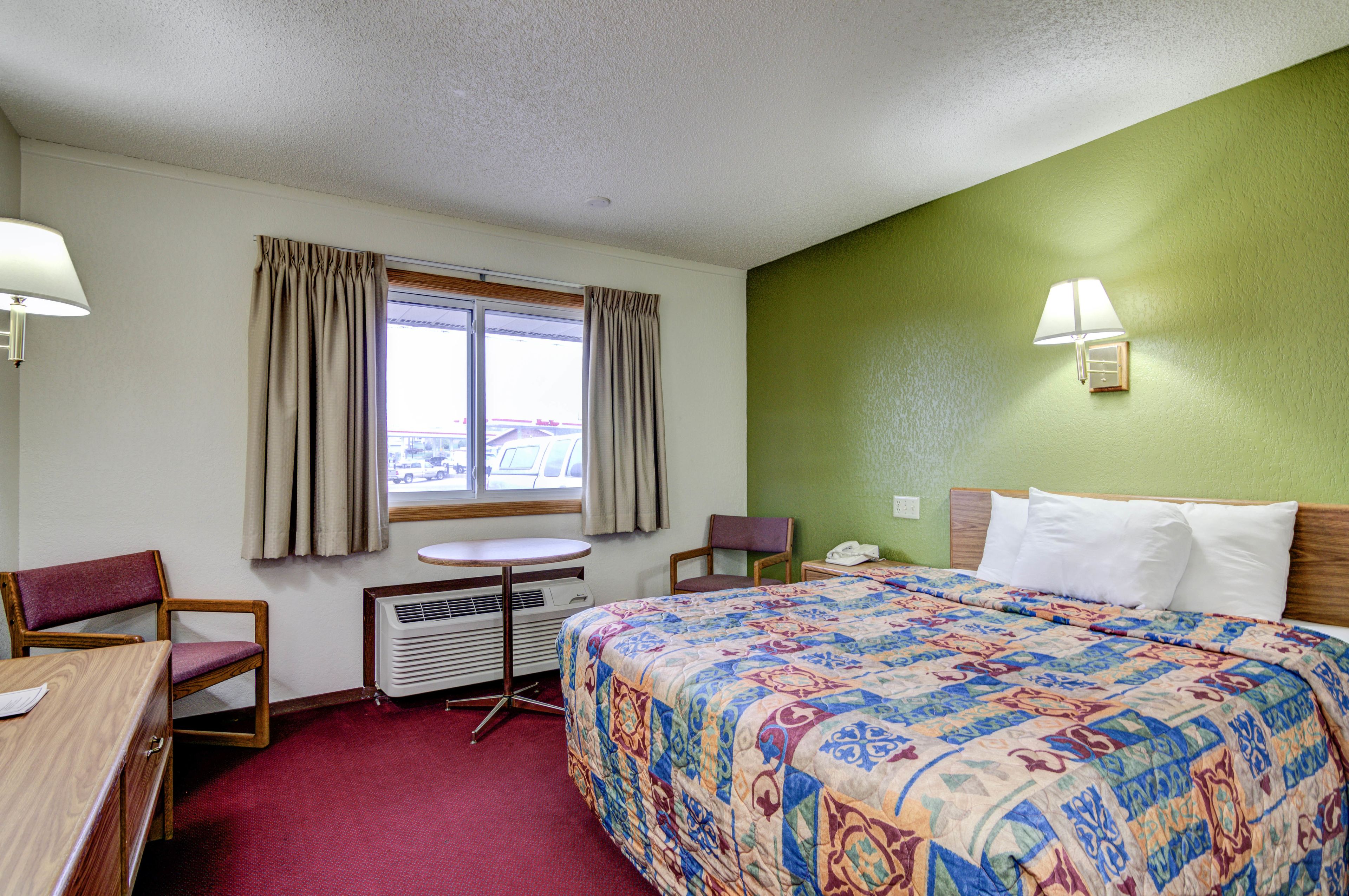Quarto de hotel com cama grande, escrivaninha, cadeira, janela com cortinas e luminária na parede.