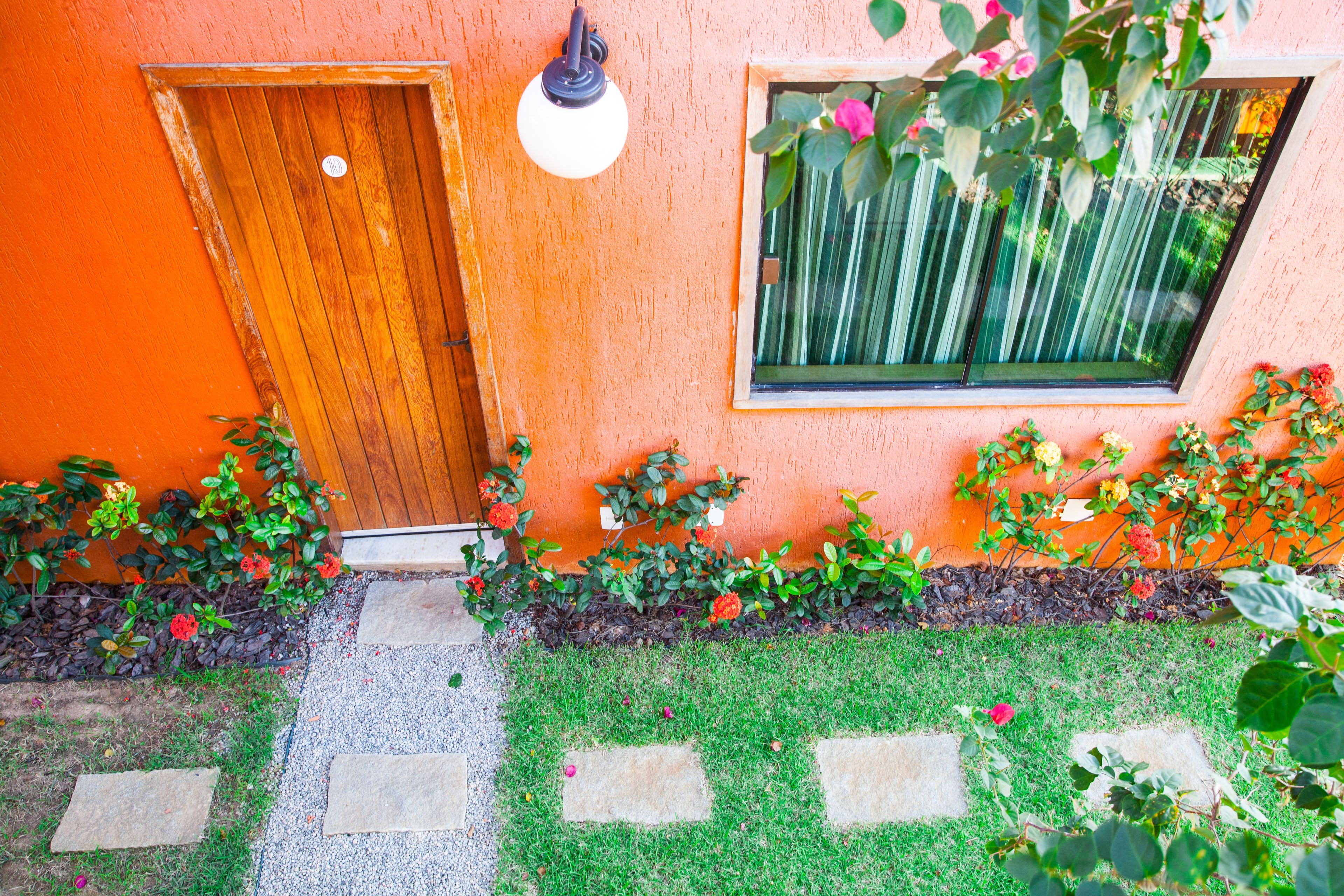 Uma casa com porta de madeira, janela com vista para bambus e um jardim com pedras no chão e flores.