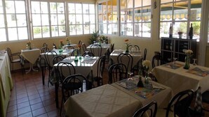 Um restaurante com várias mesas e cadeiras, janelas amplas e piso de azulejos.