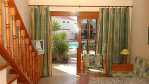 Quarto com cama, uma mesa pequena, uma cadeira, escada de madeira e vista para área de piscina externa.