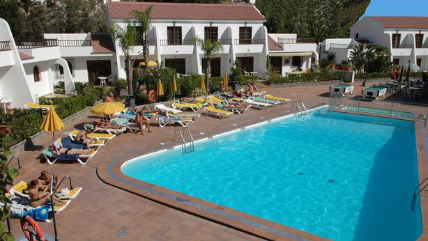 Uma área de piscina com espreguiçadeiras e guarda-sóis, cercada por edifícios brancos com telhados de telhas vermelhas.