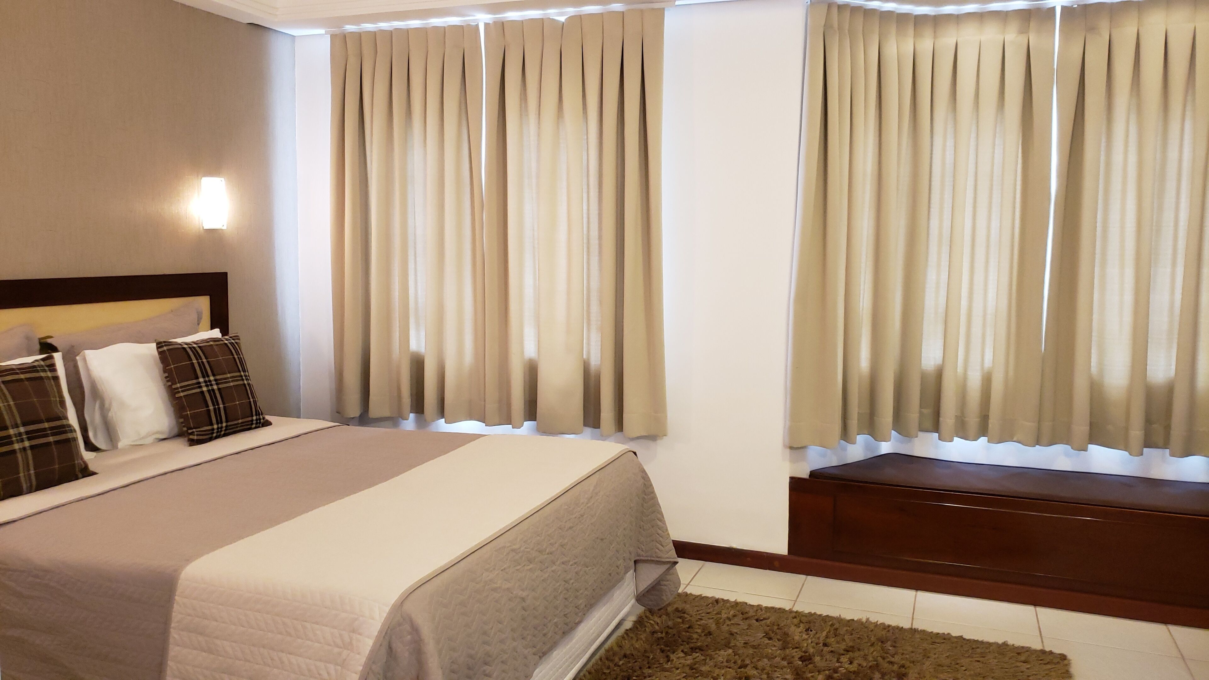 Cama bem arrumada com cabeceira, duas almofadas e um cobertor. Há duas janelas com cortinas, um banco de madeira e um tapete pequeno no chão.
