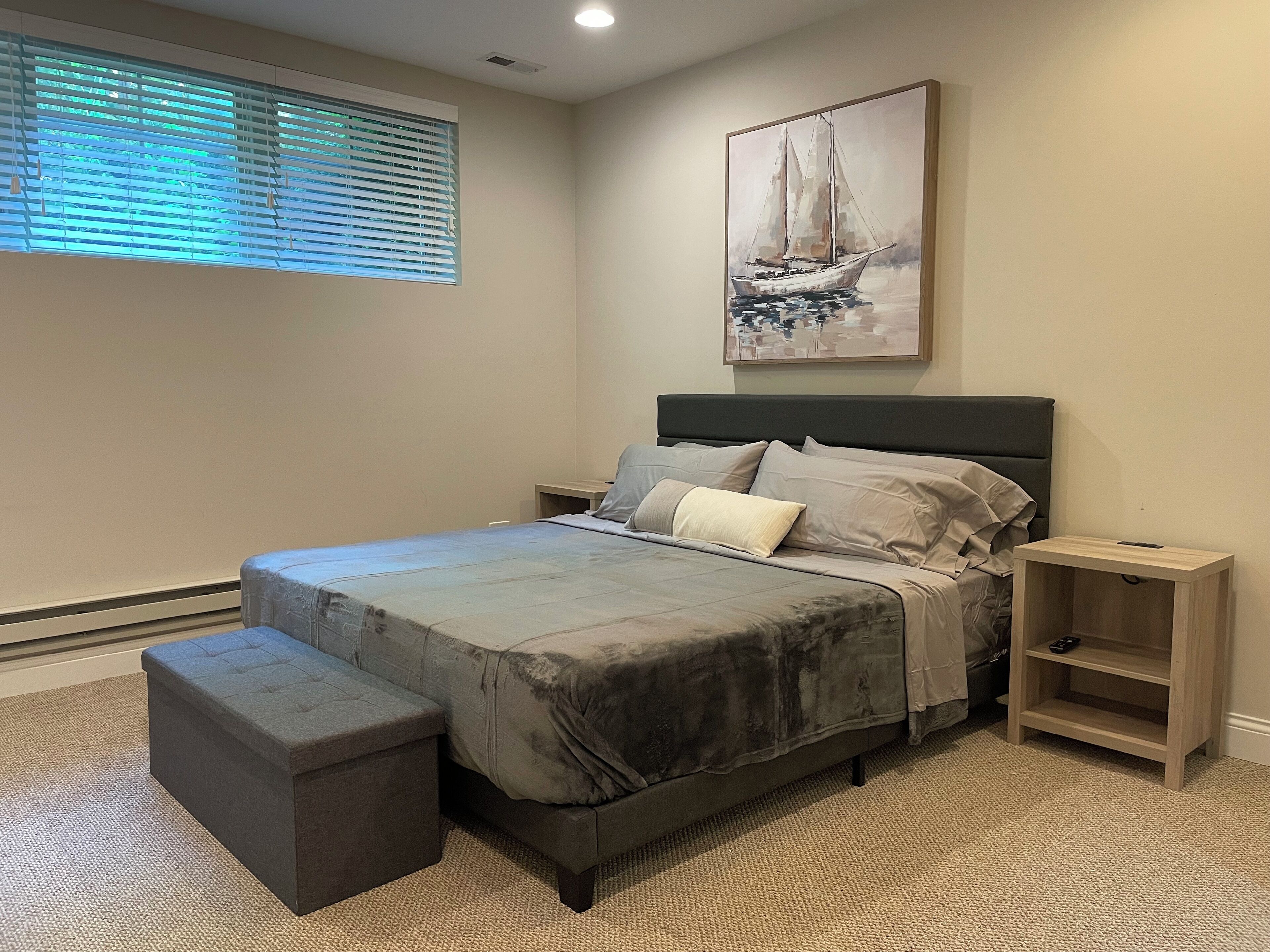 Quarto com cama, mesa de cabeceira, um banquinho e um quadro na parede.