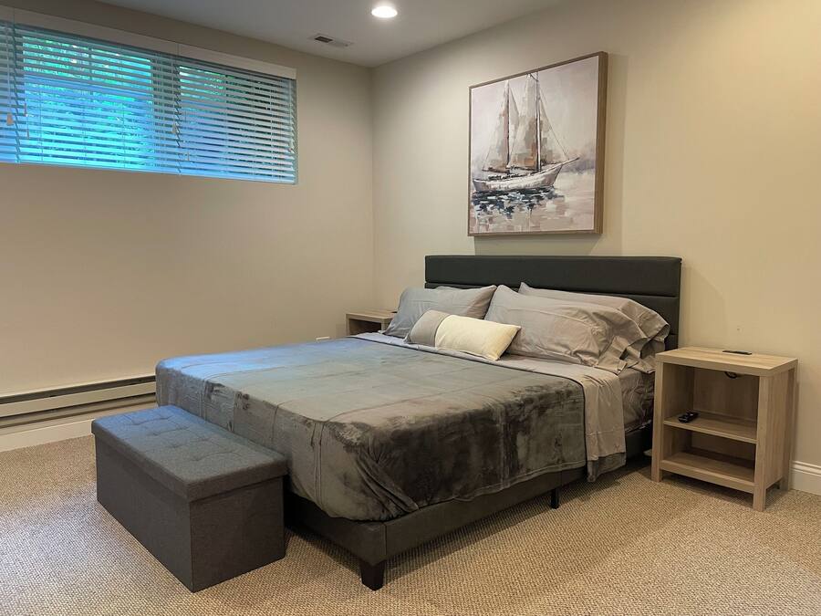 Quarto com cama, mesa de cabeceira, um banquinho e um quadro na parede.