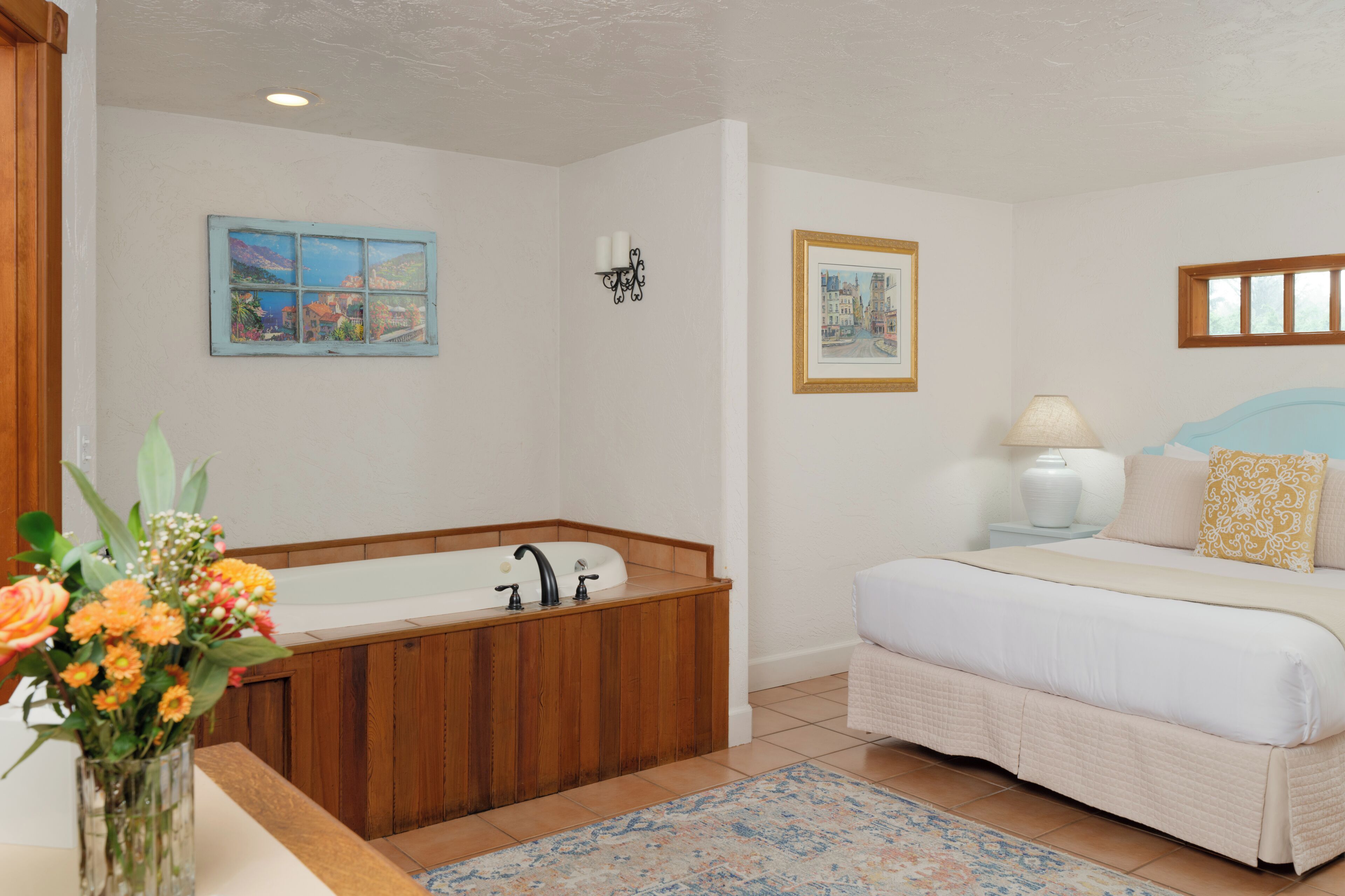 Quarto com banheira de madeira, cama com roupa de cama branca, mesa de cabeceira com abajur e duas imagens emolduradas na parede.