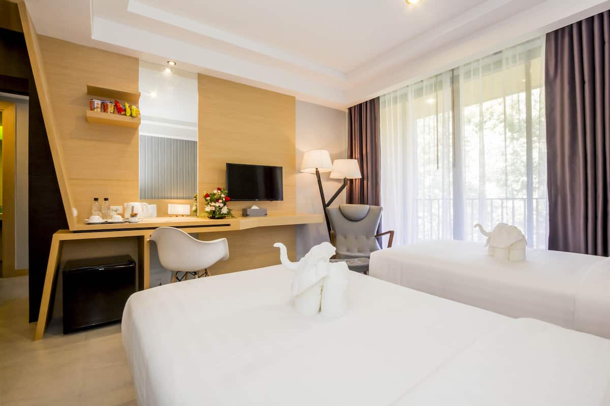 Quarto de hotel com duas camas, uma escrivaninha com televisão, uma cadeira, um abajur e uma janela com cortinas.