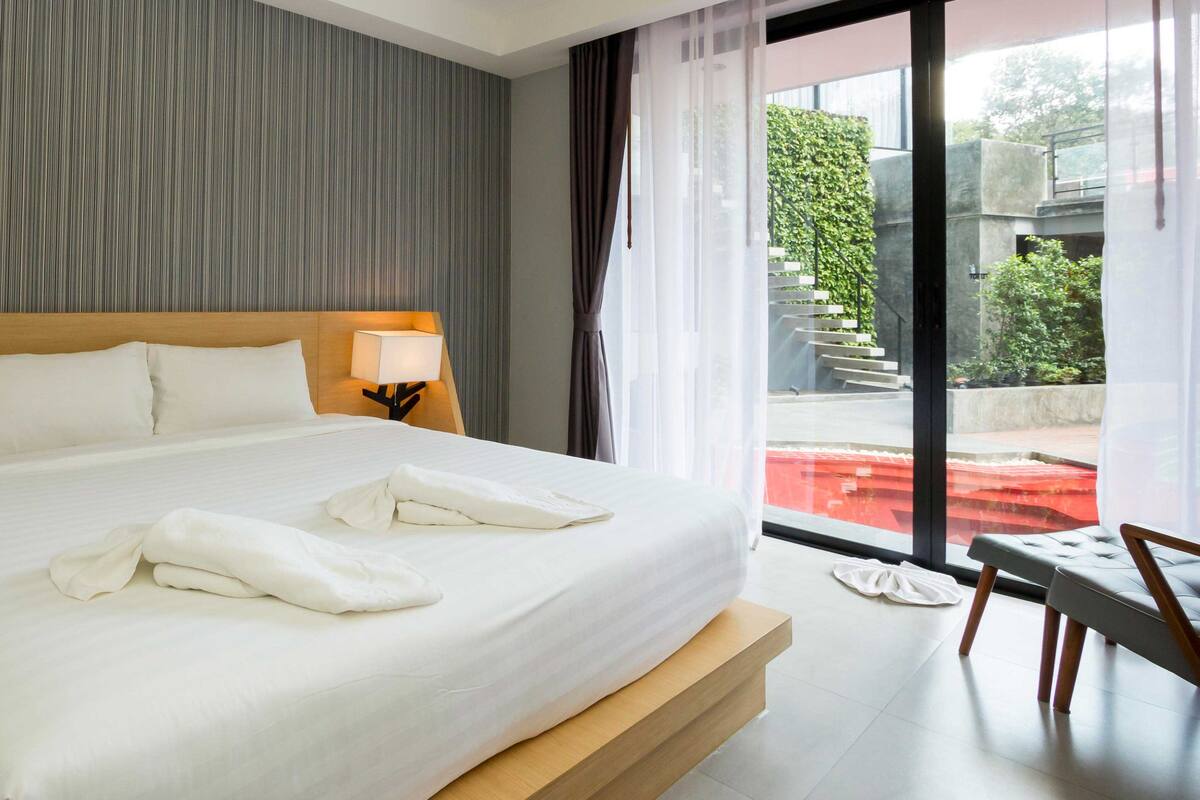 Quarto de hotel moderno com uma cama grande, duas mesas de cabeceira com luminárias, vista para uma piscina vermelha e uma varanda com vegetação.