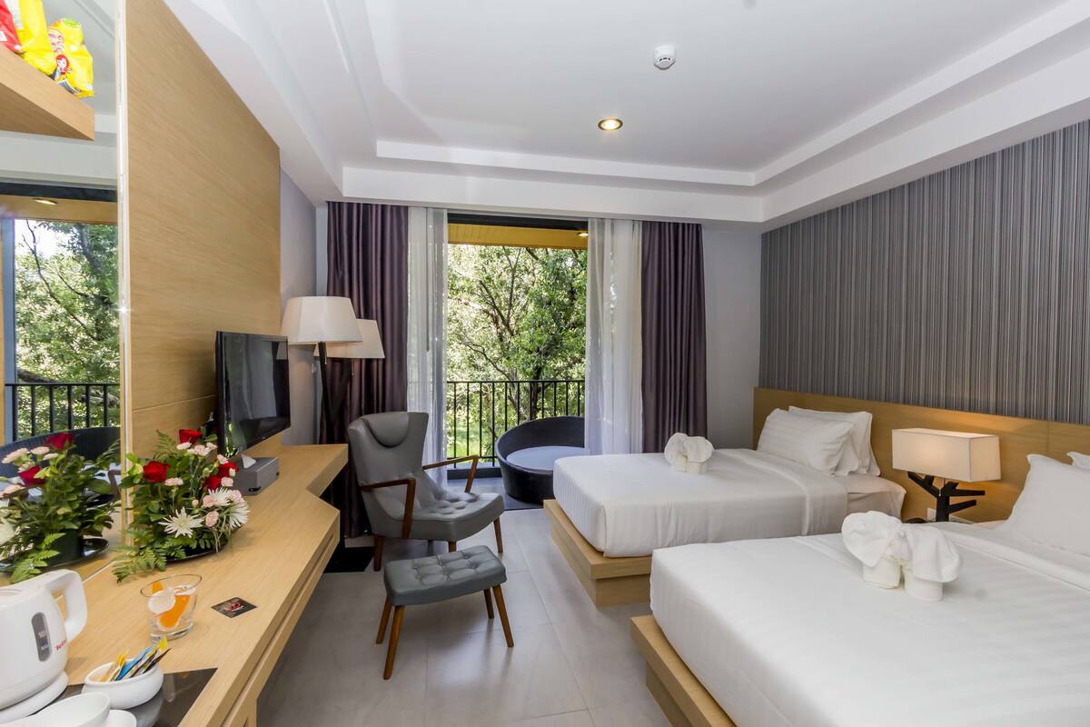 Quarto de hotel com duas camas, uma mesa de trabalho com televisão, uma cadeira, uma mesinha com arranjo floral e vista para árvores pela janela.