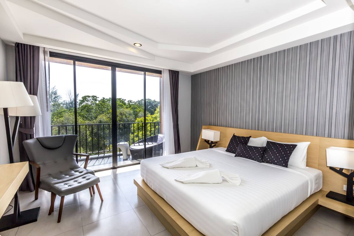 Quarto de hotel moderno com uma cama grande, uma cadeira, uma mesa de trabalho e uma varanda com vista para área verde.